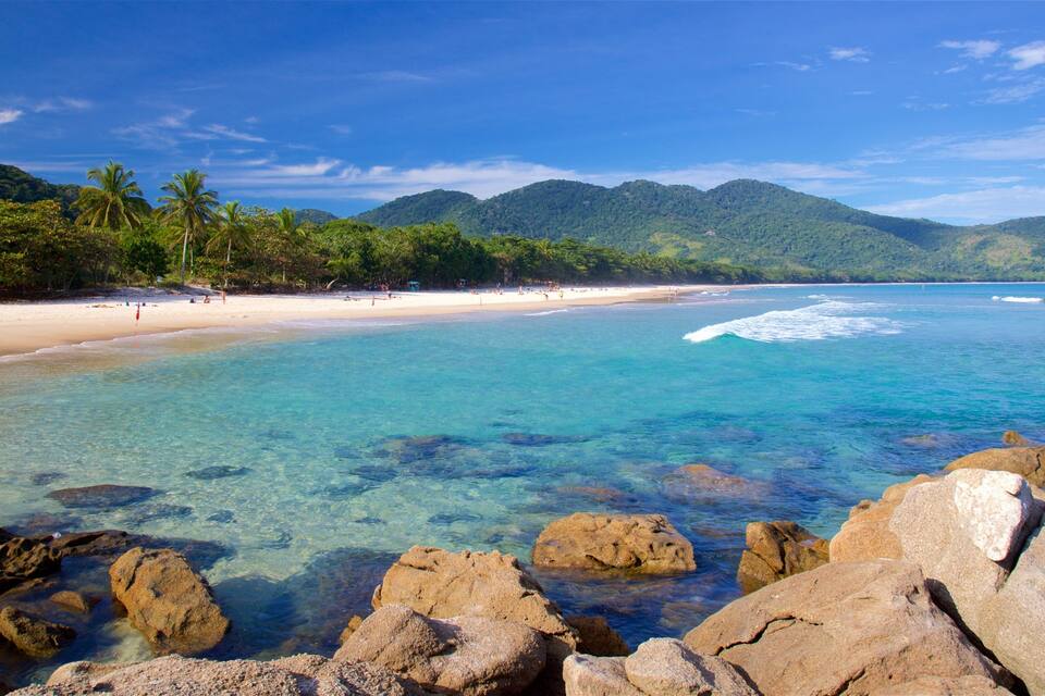 Uma praia com águas turquesa cristalinas, um primeiro plano rochoso e colinas verdejantes ao fundo.