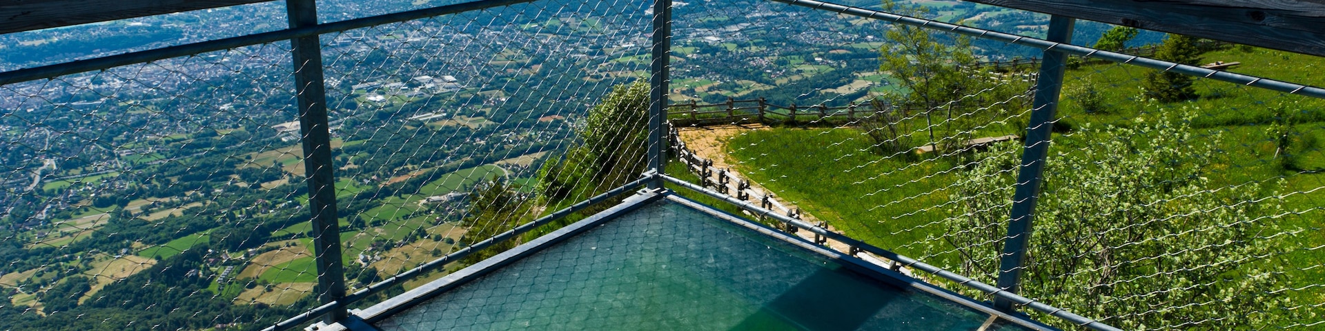 Plataforma panorâmica com fundo de vidro e guarda-corrimão de metal, com vista para um vale e montanhas ao longe.