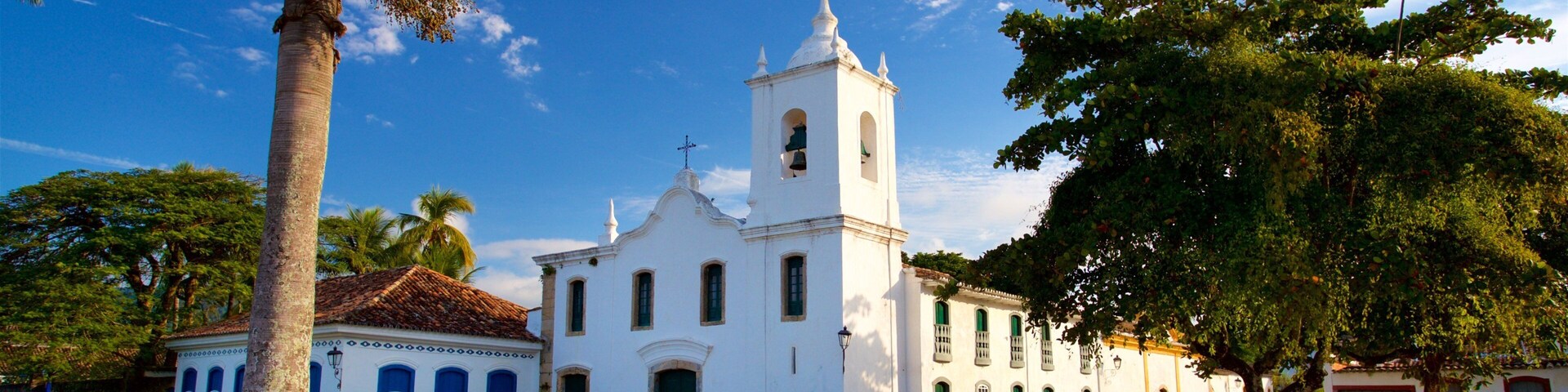 Uma igreja branca com um campanário, rodeada por palmeiras e um céu azul límpido.