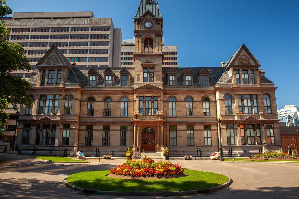 Um edifício histórico com uma torre do relógio, rodeado por um jardim bem cuidado e arranha-céus modernos ao fundo.