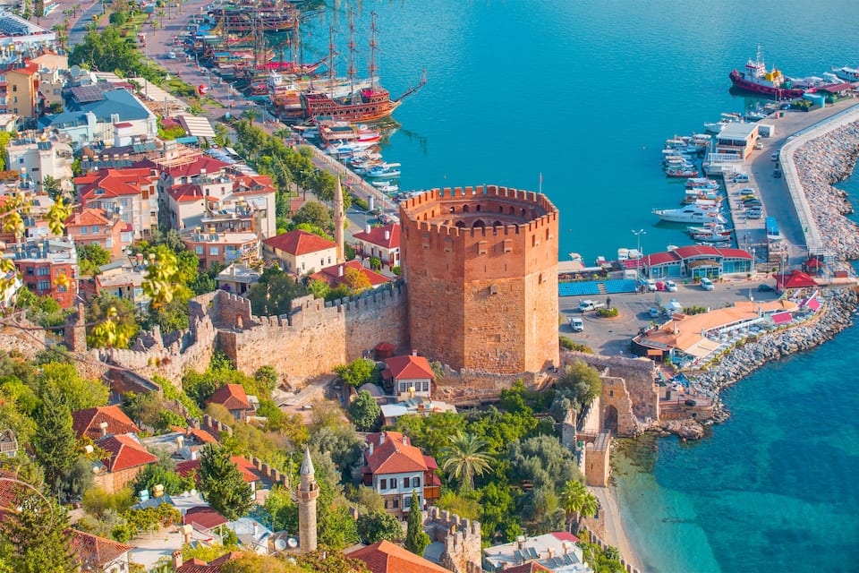 Uma cidade costeira com um castelo imponente, uma marina com barcos e uma praia.