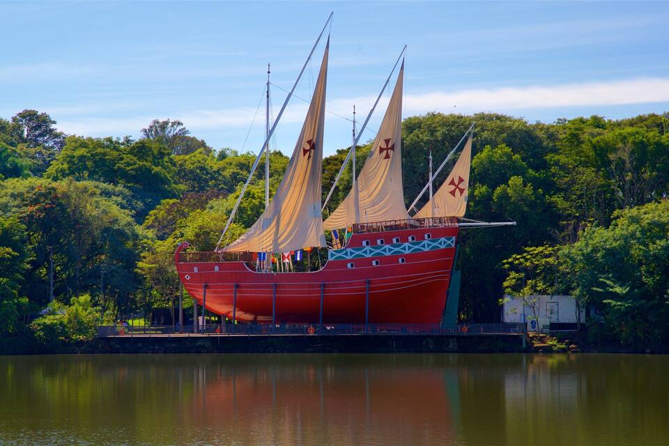 Um grande navio vermelho com velas, atracado em um lago rodeado por árvores verdes e densas.