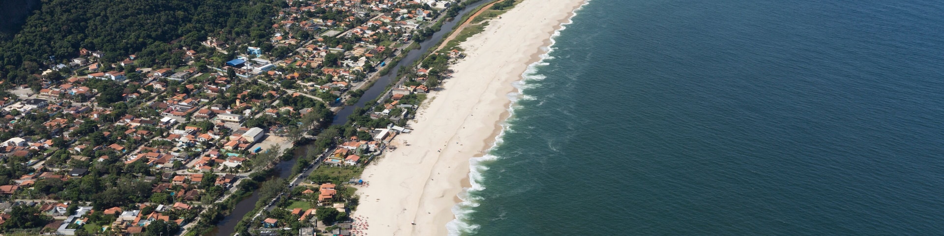 Vista aérea de uma cidade costeira com uma extensa praia, área urbana densa e colinas ao redor.