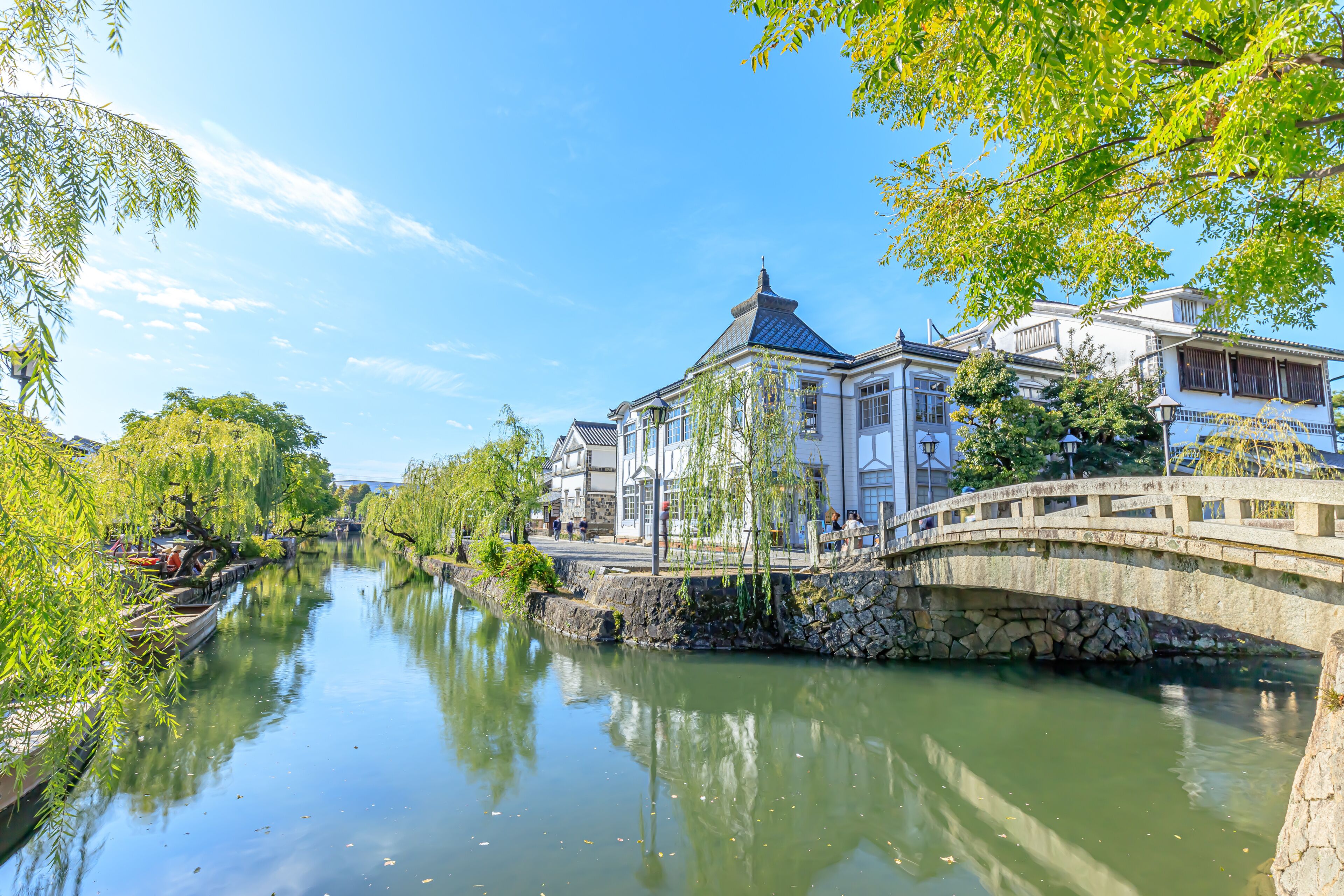 Um rio calmo com uma ponte de pedra, edifícios tradicionais e vegetação exuberante.