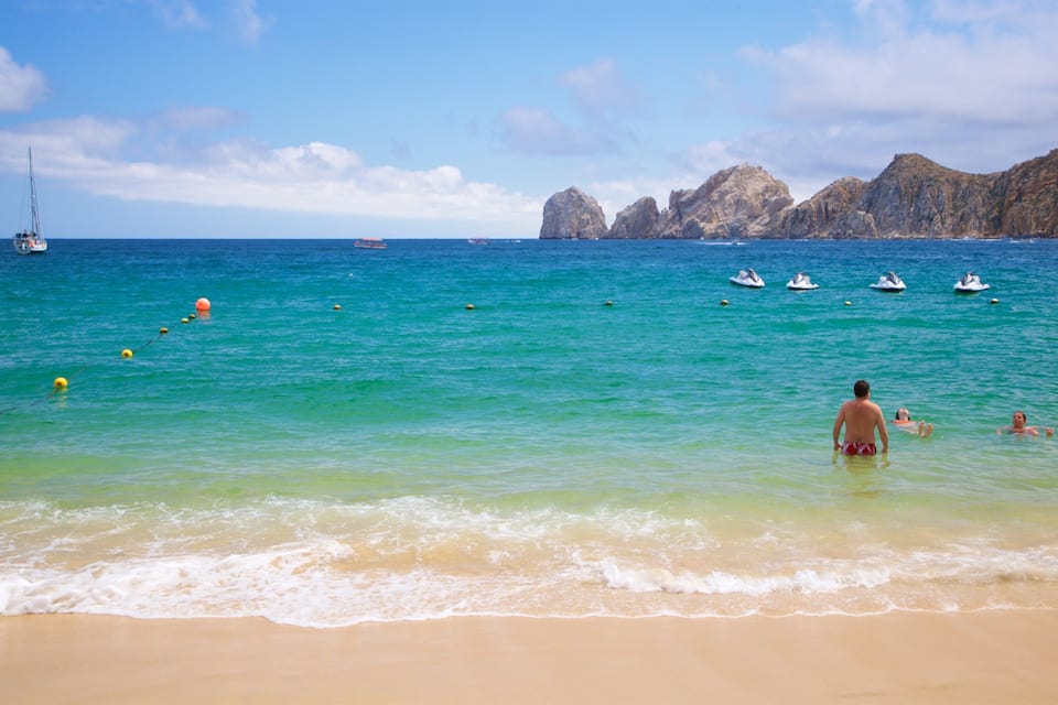 Uma praia com águas turquesa cristalinas, um veleiro e falésias rochosas ao fundo.