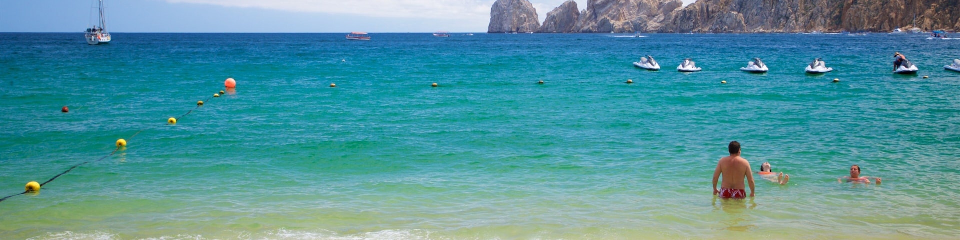 Uma praia com águas turquesa cristalinas, um veleiro e falésias rochosas ao fundo.