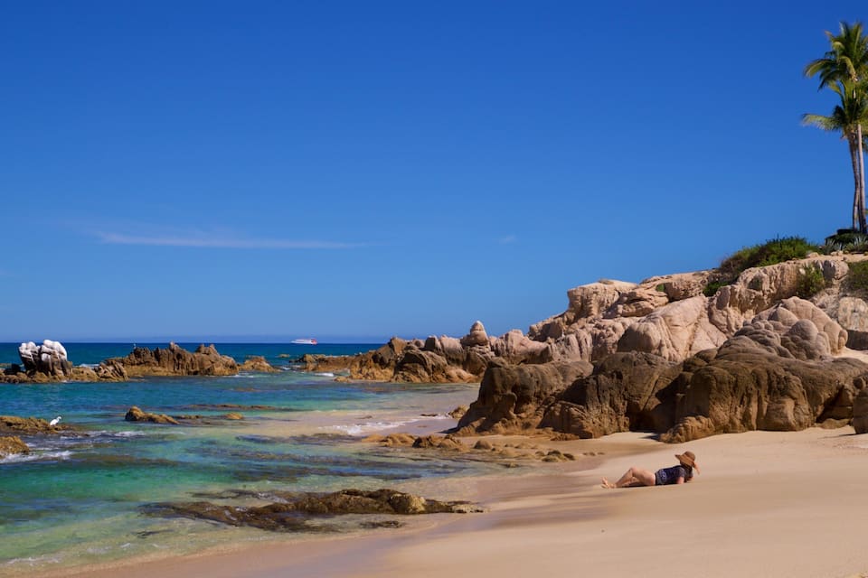 Uma praia arenosa com águas cristalinas azul-turquesa, formações rochosas e palmeiras.