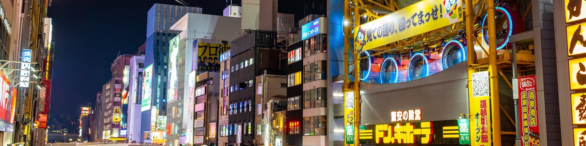 Rua movimentada à noite, com letreiros iluminados e um rio em primeiro plano.