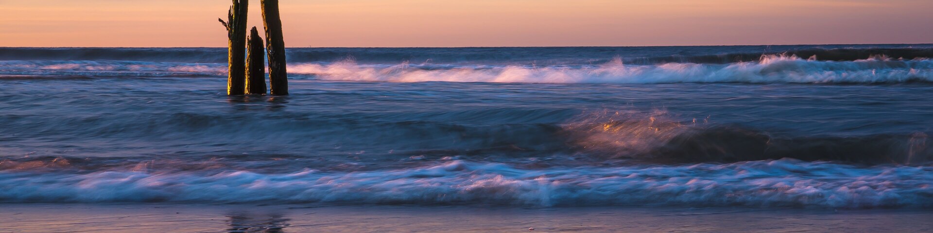 Um pôr do sol sobre o oceano, com ondas quebrando na praia e duas estacas de madeira na água.