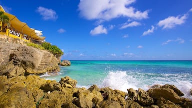 Costa rochosa com águas turquesa cristalinas, um edifício amarelo com varanda voltada para o mar e um céu azul límpido com nuvens esparsas.