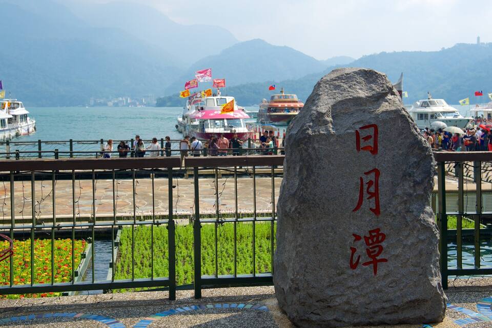 Uma pedra com caracteres chineses, um cais com barcos e montanhas ao fundo.
