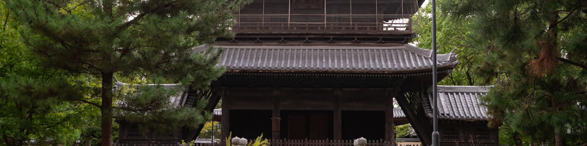 Um edifício japonês tradicional com telhado de telhas e estrutura de madeira, cercado por árvores e um caminho de pedra.