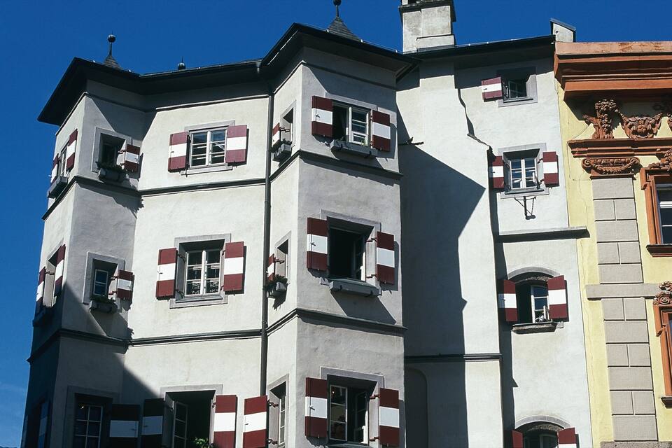 Um edifício de vários andares com portadas vermelhas e brancas, chaminé e cornija decorativa.