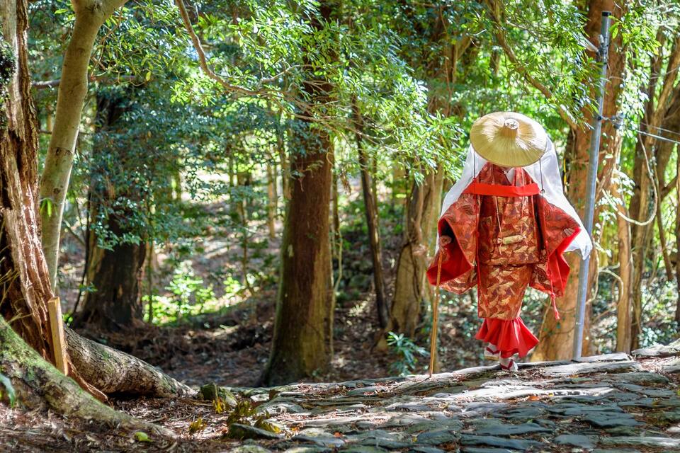 Uma trilha na floresta com uma pessoa vestida com traje tradicional caminhando à distância.