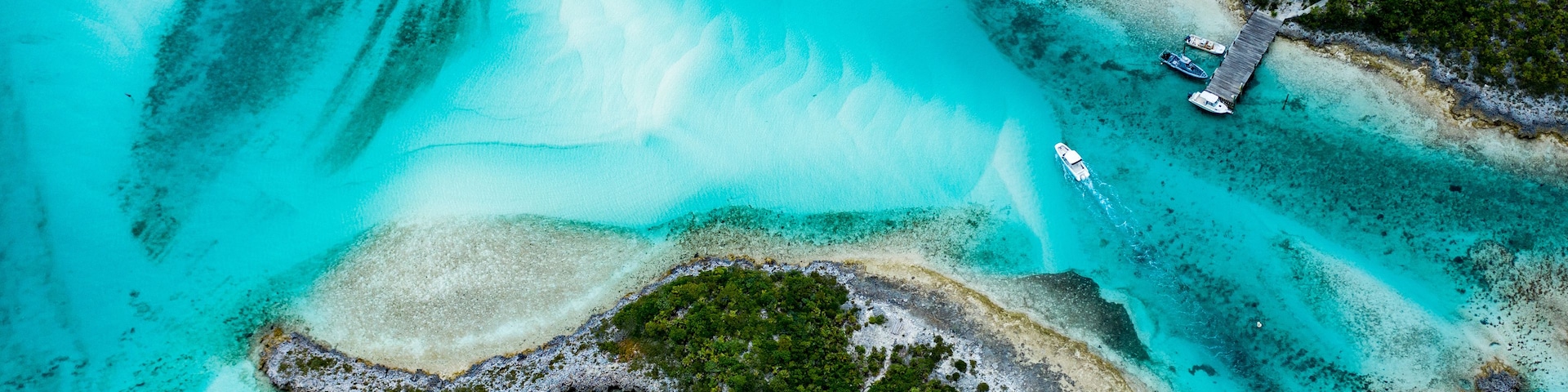 Vista aérea de uma ilha tropical com águas turquesa cristalinas, um pequeno píer e alguns edifícios cercados por vegetação exuberante.