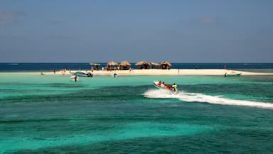 Uma praia tropical com águas turquesa cristalinas, um barco a grande velocidade na água e palafitas no fundo.