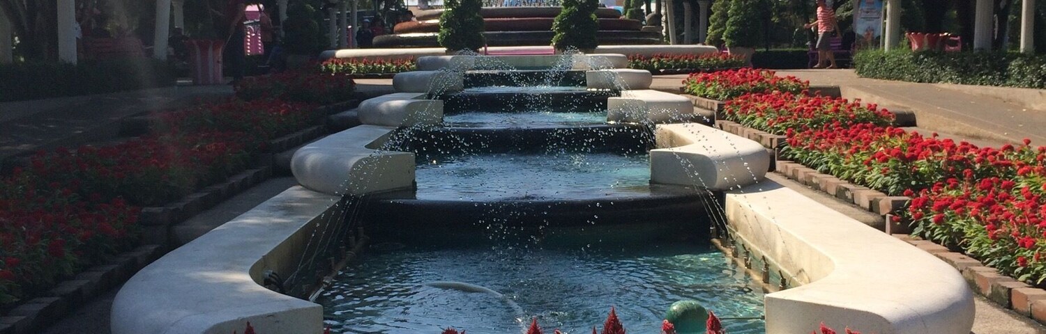 Um jardim com uma fonte central, rodeado por flores vermelhas e sebes bem aparadas.