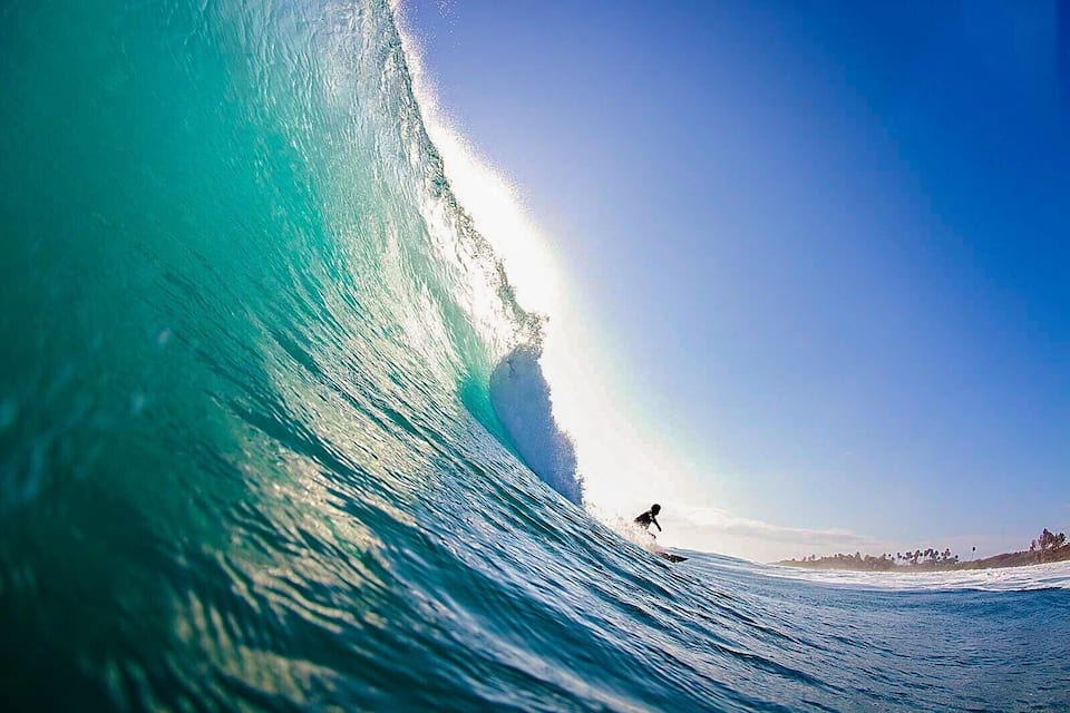 Um surfista desliza sobre uma onda grande, com água azul cristalina e céu ensolarado.