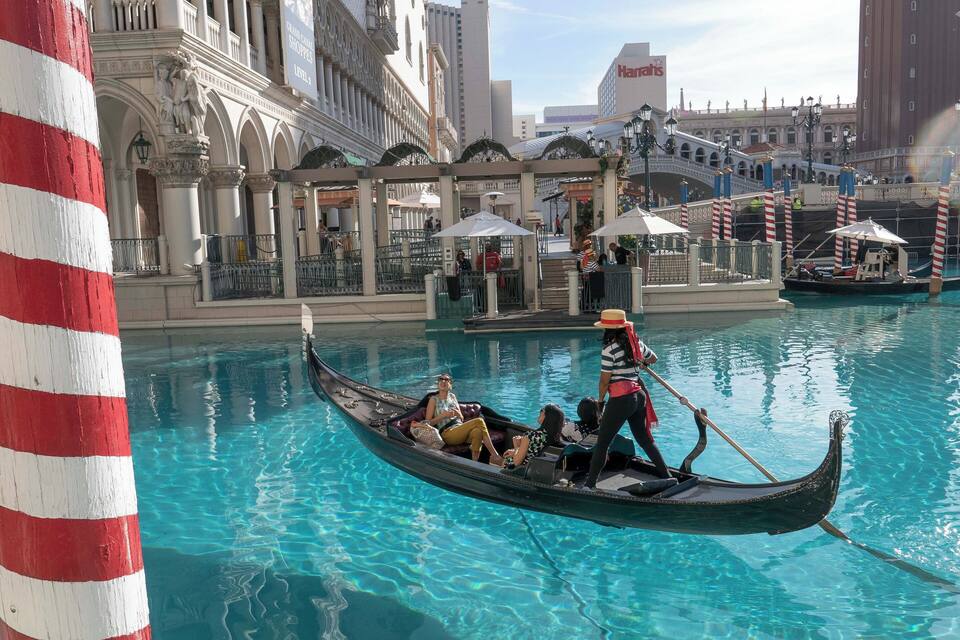Uma gôndola num canal com turistas, um mastro vermelho e branco, e edifícios históricos ao fundo.