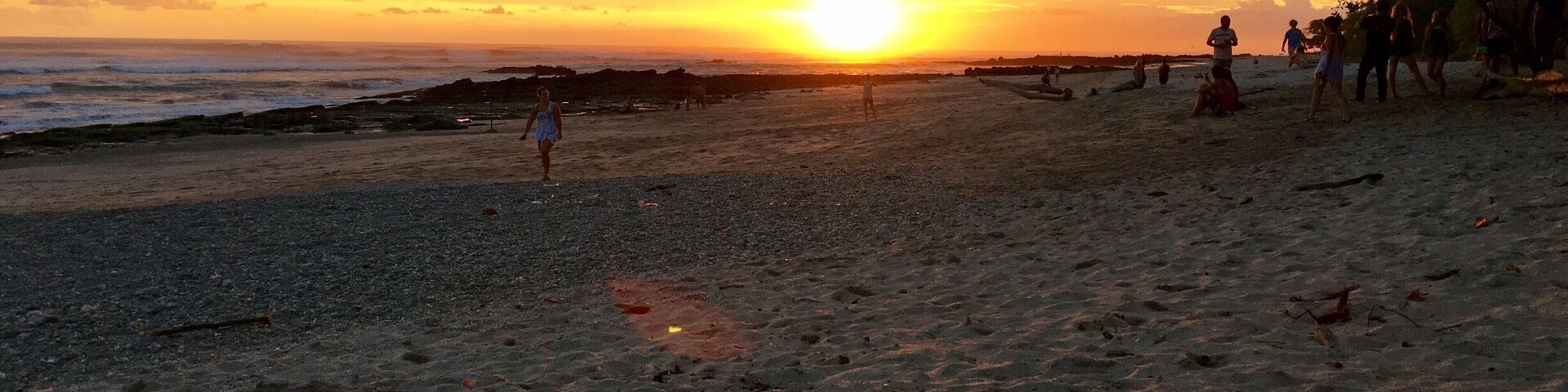 Uma praia ao pôr do sol, com o sol baixo no horizonte, pessoas caminhando pela orla e um afloramento rochoso ao fundo.