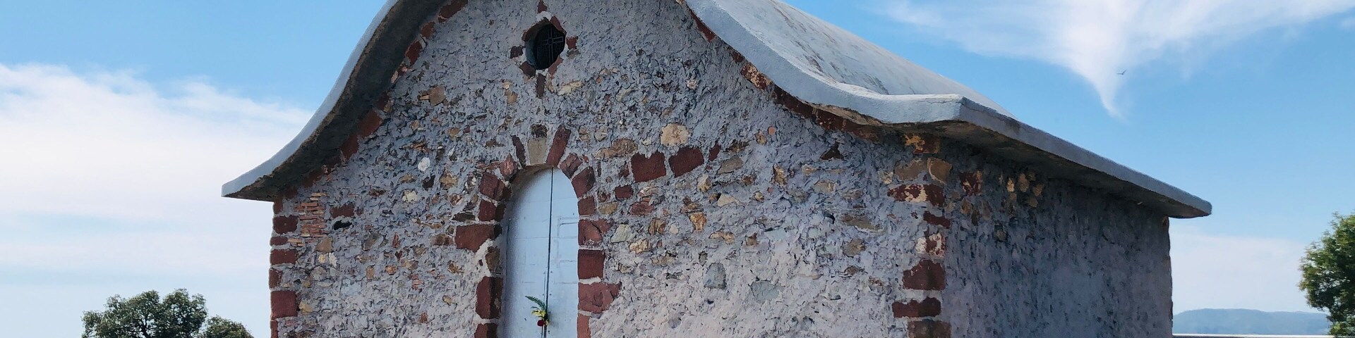 Uma pequena igreja desgastada pelo tempo com uma cruz no topo, uma bicicleta estacionada na frente e um céu azul claro ao fundo.