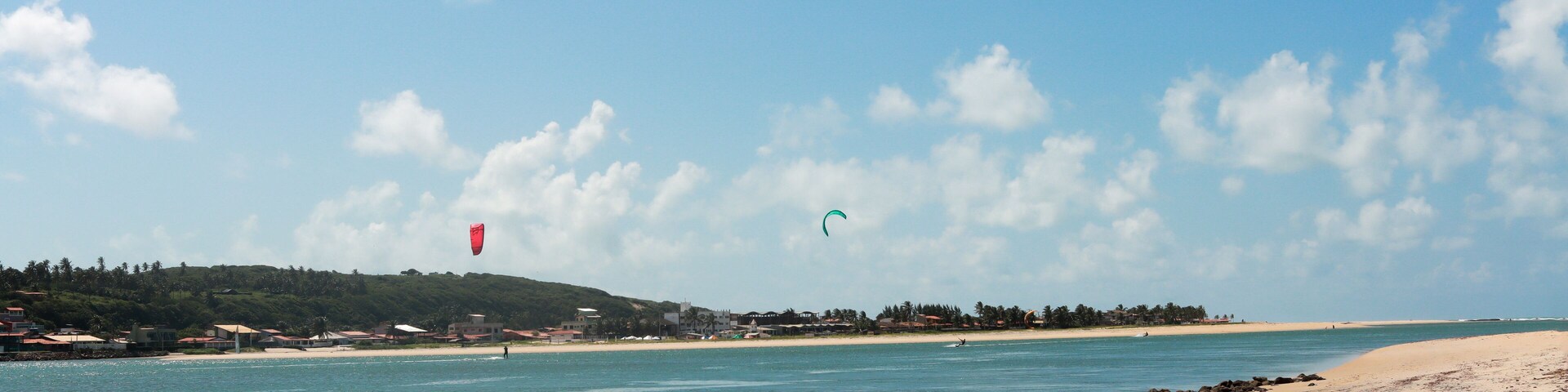 Uma praia com águas azul-claras, uma costa rochosa e uma colina distante com edifícios.