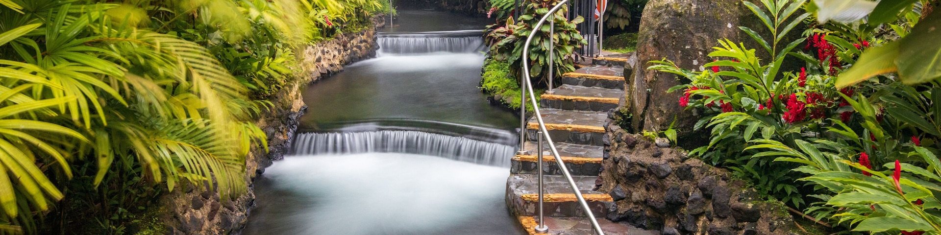 Uma cascata despencando sobre um afloramento rochoso, cercada por vegetação tropical exuberante e uma estrutura com telhado de palha.