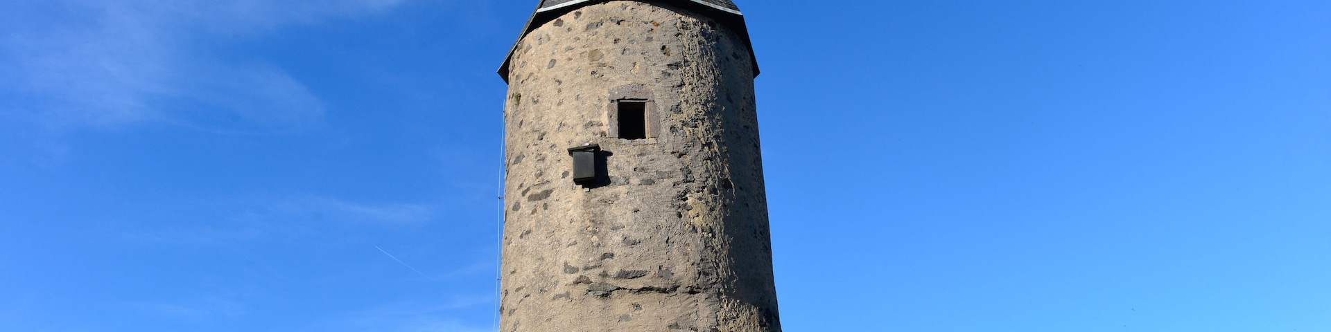 Uma torre de pedra alta e cilíndrica com telhado cónico, uma pequena janela e uma porta, rodeada por uma muralha de pedra e um guarda-corpo de metal.