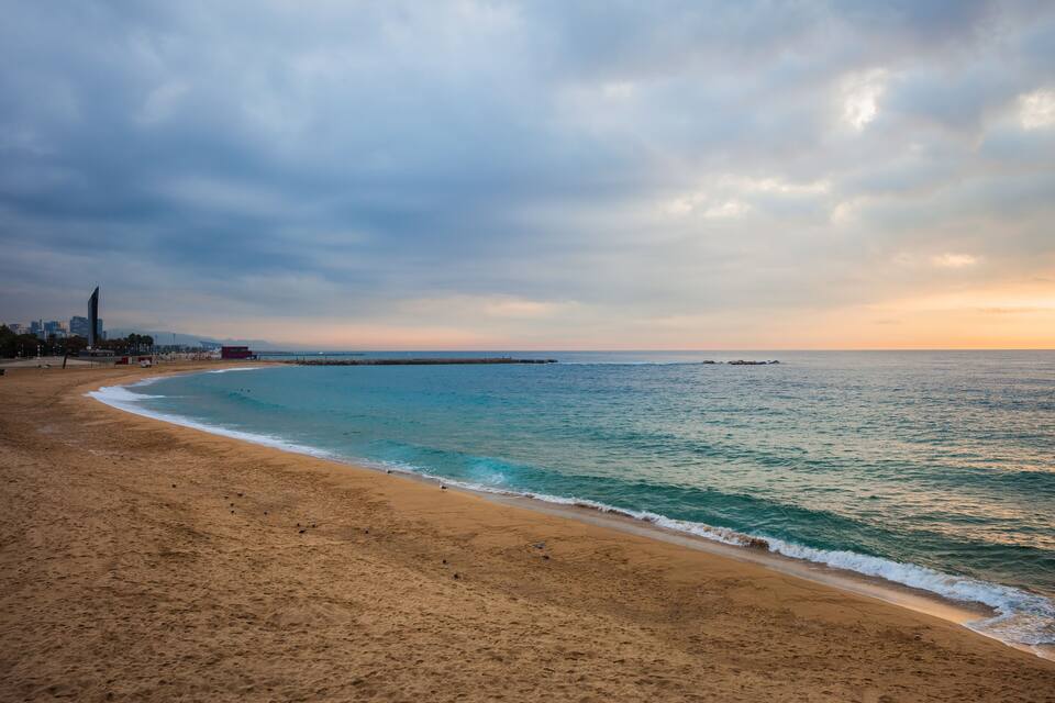 Uma praia com costa sinuosa, um horizonte urbano distante e céu nublado.