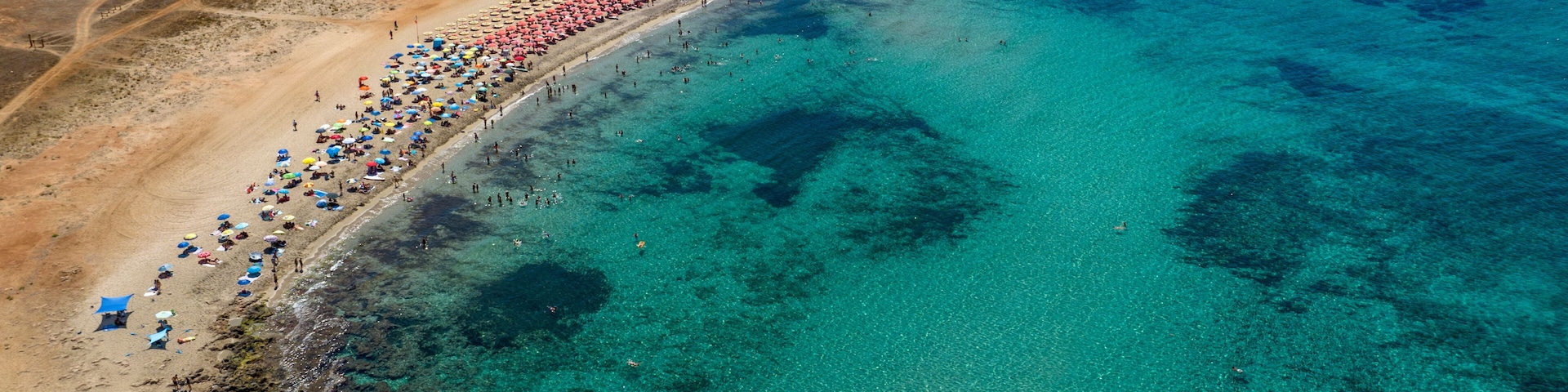 Vista aérea de uma praia com águas turquesa cristalinas, uma extensa fileira de guarda-sóis e um litoral rochoso.