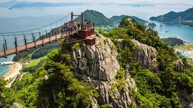 Uma ponte suspensa com pessoas caminhando, com vista para uma área costeira com montanhas e céu claro.