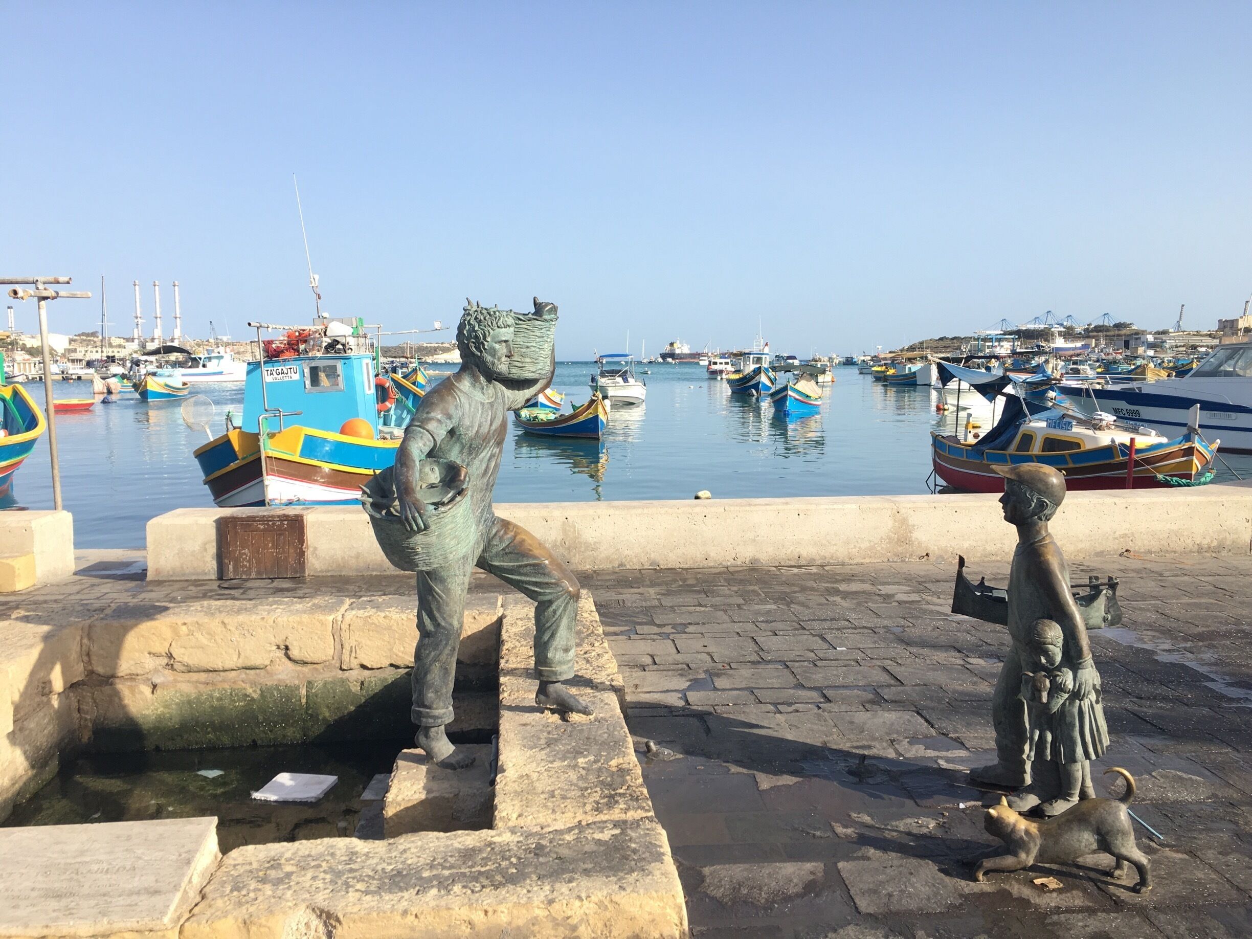 Um porto com barcos, uma estátua de um homem com um peixe e outra estátua de uma mulher com um cão.