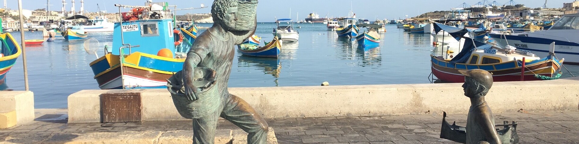 Um porto com barcos, uma estátua de um homem com um peixe e outra estátua de uma mulher com um cão.
