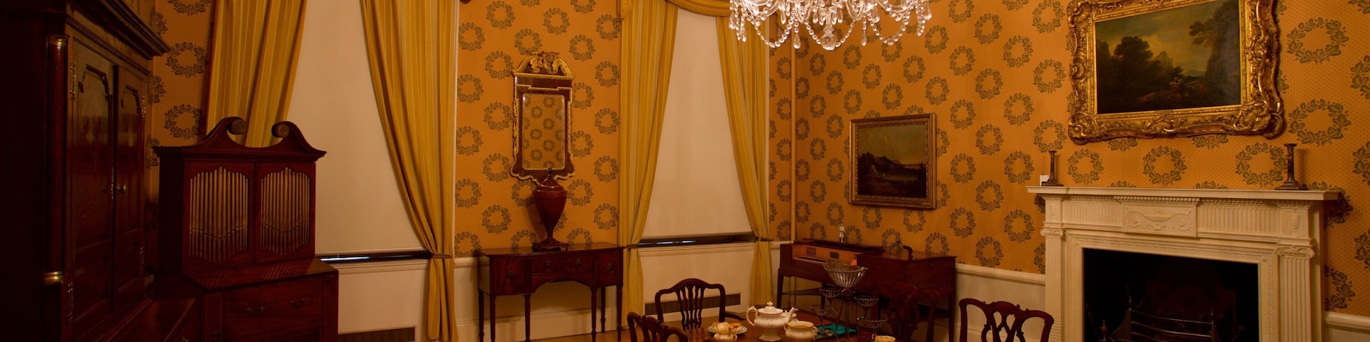 Uma sala de jantar com uma mesa de madeira posta para refeição, um candelabro, lareira e papel de parede estampado.