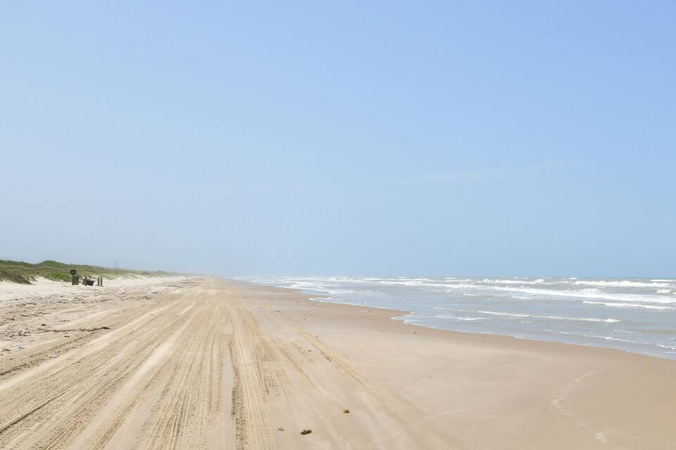 Uma praia de areia com ondas quebrando na costa e dunas ao fundo.