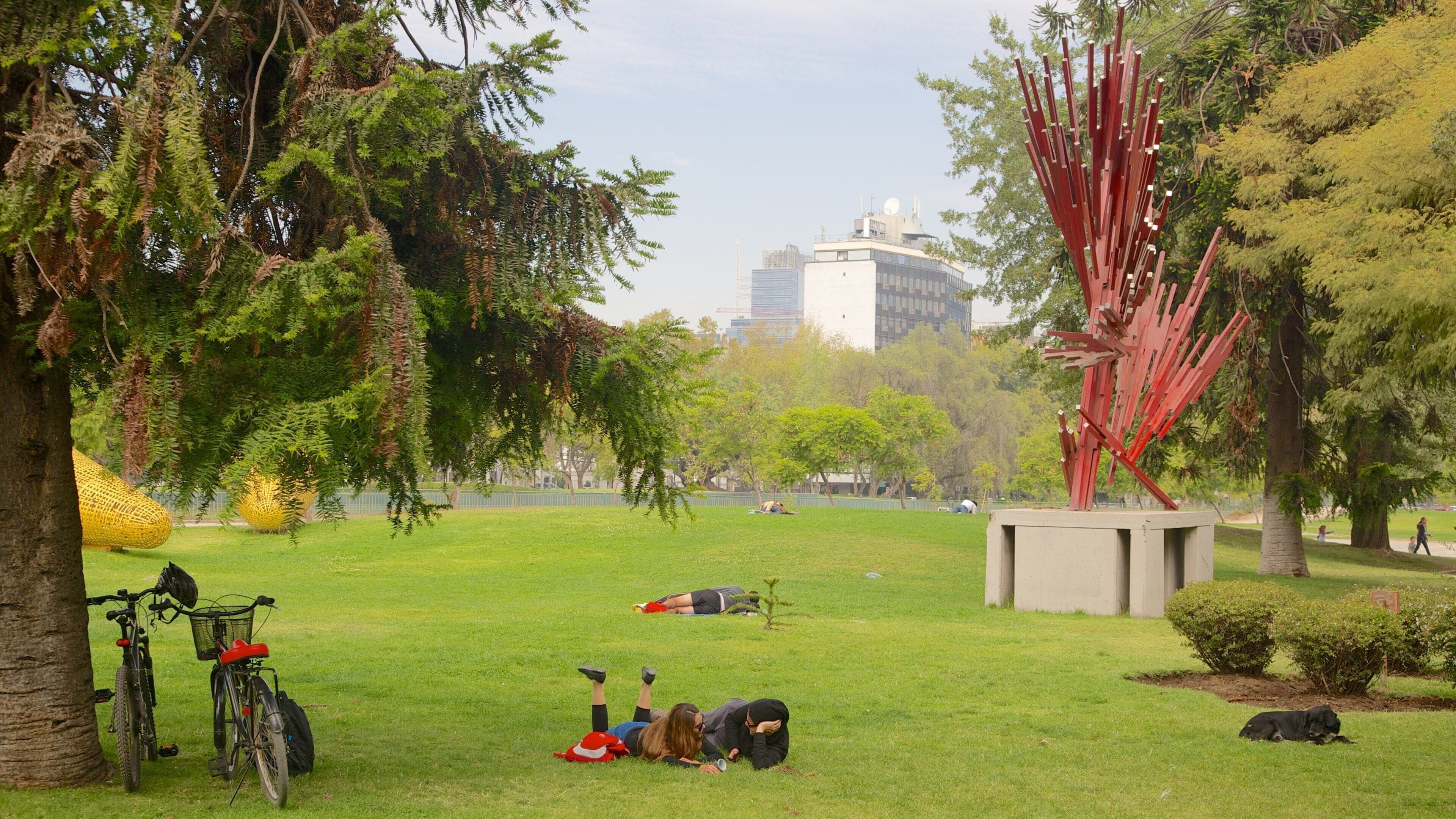 Um parque com grama verde, árvores, uma escultura vermelha e uma bicicleta.