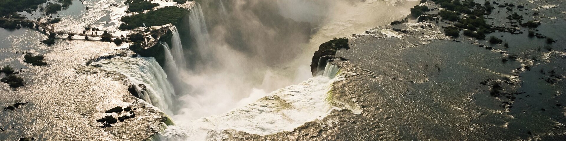 Uma grande cascata com névoa subindo da base, cercada por água e vegetação.