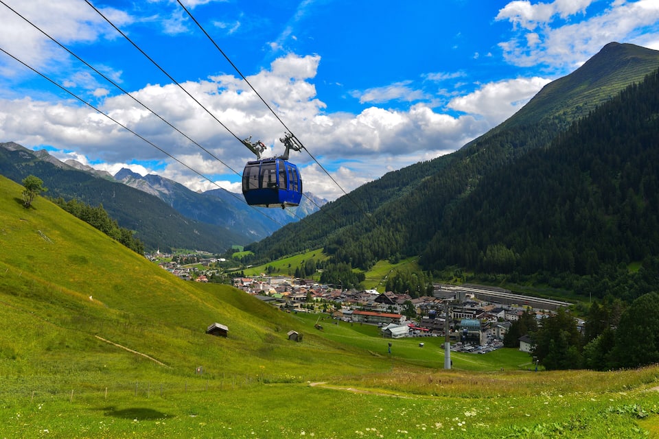 Um teleférico movendo-se sobre um vale, com uma cidade abaixo, cercada por montanhas.