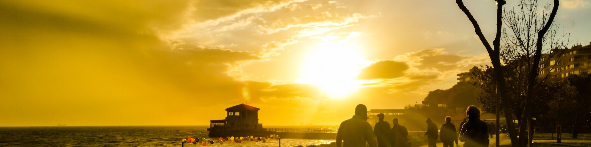 Um pôr do sol sobre uma área costeira com um cais, pessoas caminhando e um edifício na orla.