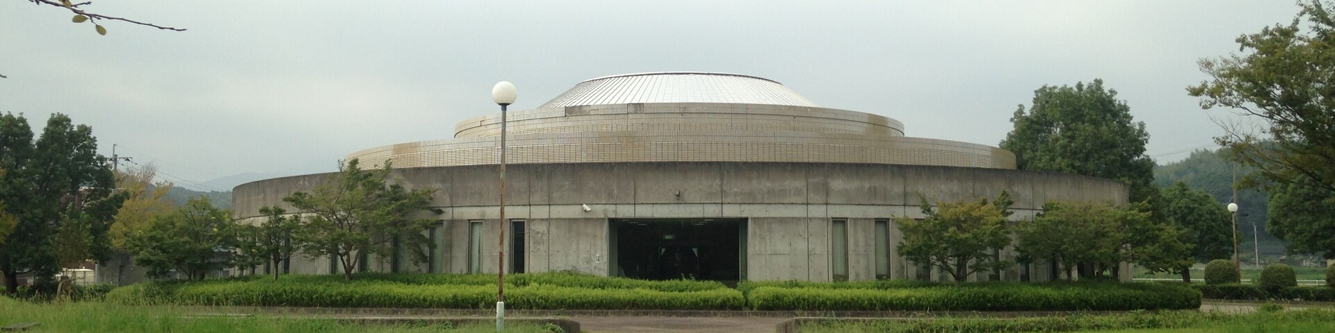 Um edifício grande e circular com cúpula, rodeado por um caminho pavimentado e vegetação.