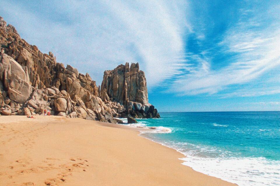 Uma praia arenosa com um afloramento rochoso e céu azul límpido.
