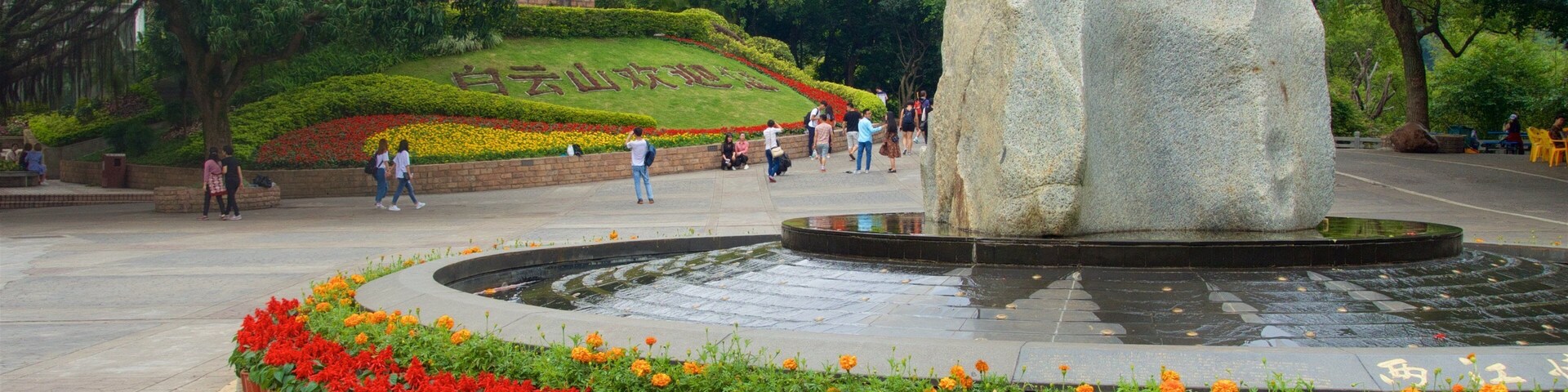 Um parque com uma grande escultura de pedra, uma fonte e canteiros de flores.
