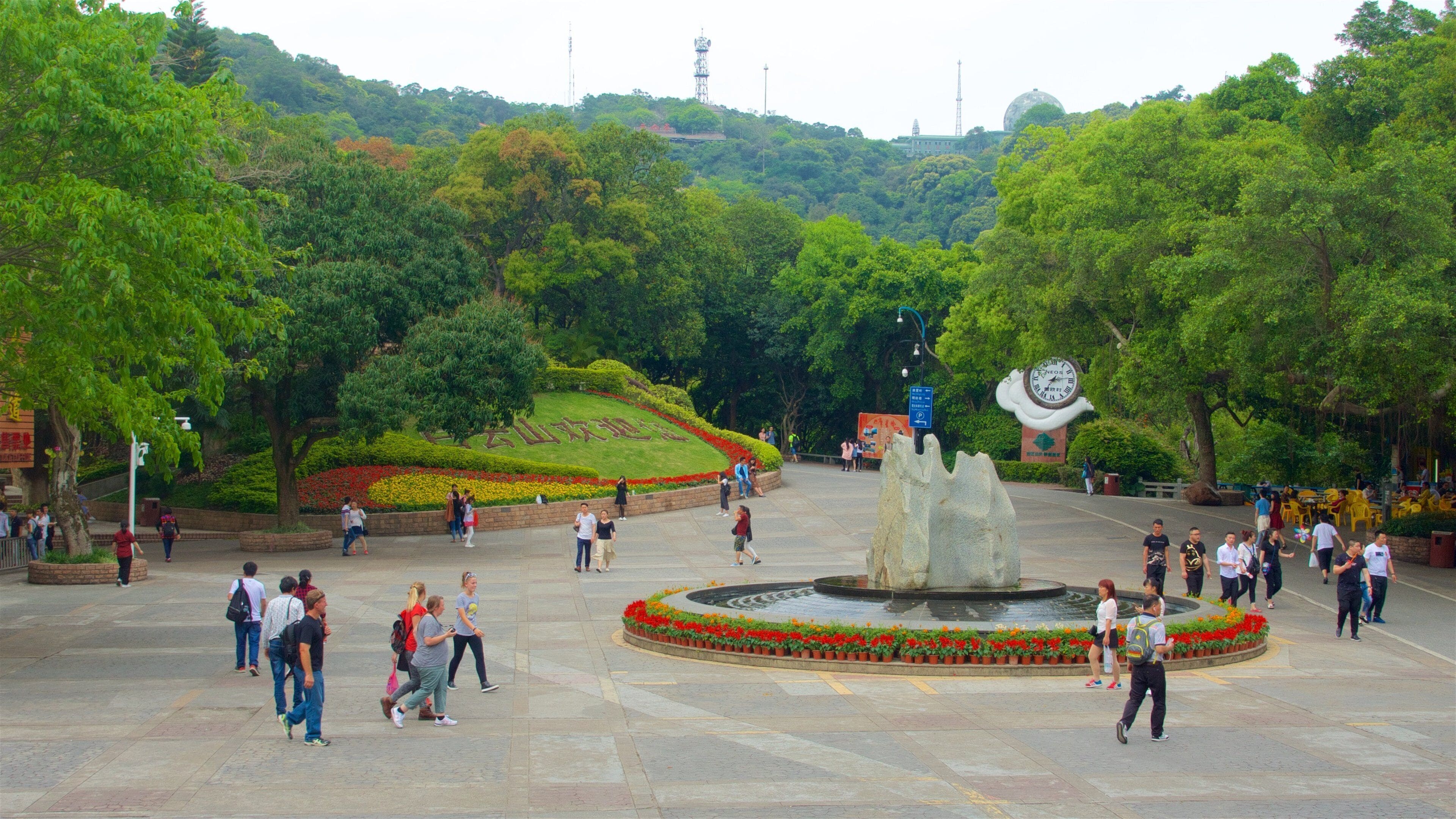 Uma praça pública com uma fonte central, rodeada por vegetação exuberante e uma colina ao fundo.