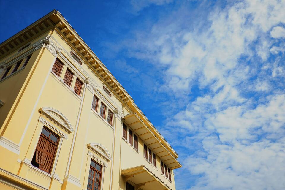 Um edifício amarelo com persianas castanhas e um céu azul com nuvens.