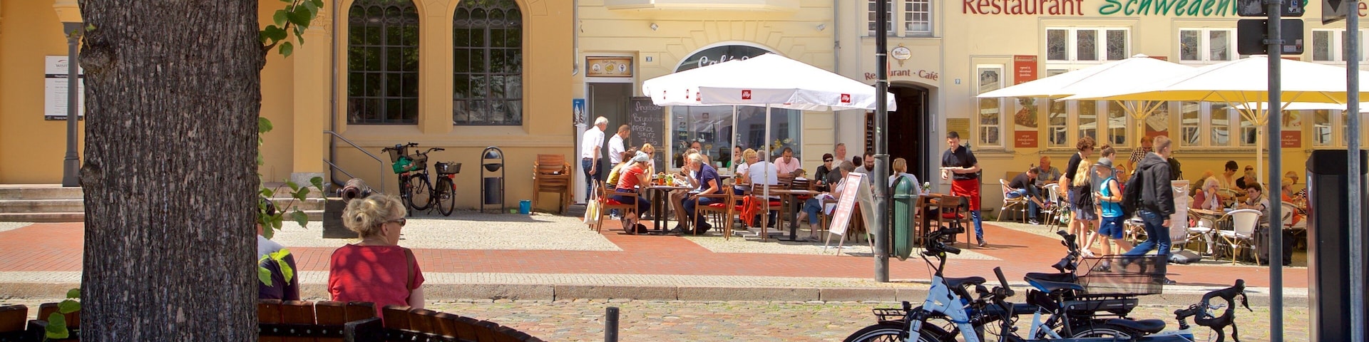 Uma praça de paralelepípedos com mesas ao ar livre em cafés, bicicletas e uma árvore.