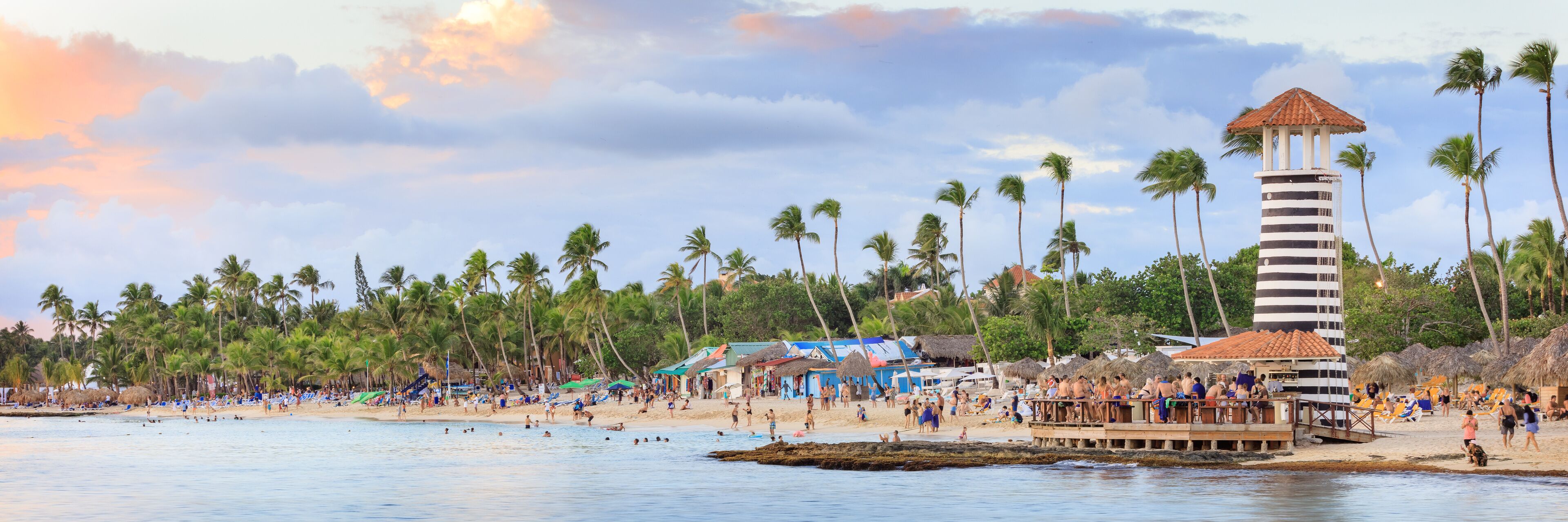 Uma praia com palmeiras, uma torre salva-vidas e pessoas.