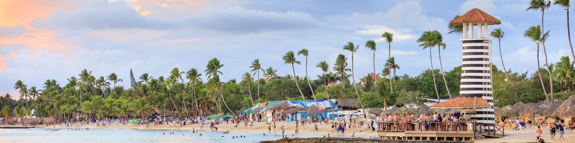 Uma praia com palmeiras, uma torre salva-vidas e pessoas.