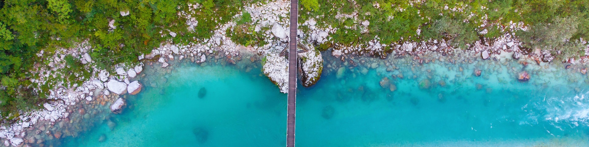 Um rio azul-turquesa com uma ponte de madeira, cercado por uma floresta verde densa.