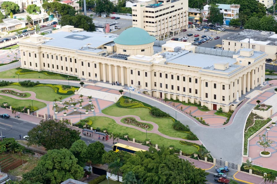 Vista aérea de um edifício grande com cúpula, rodeado por jardins ajardinados e uma estrada curva.
