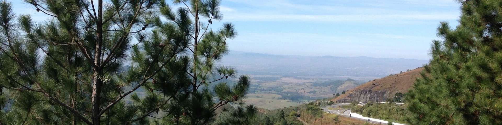 Paisagem montanhosa com pinheiros densos e uma estrada sinuosa ao longe.